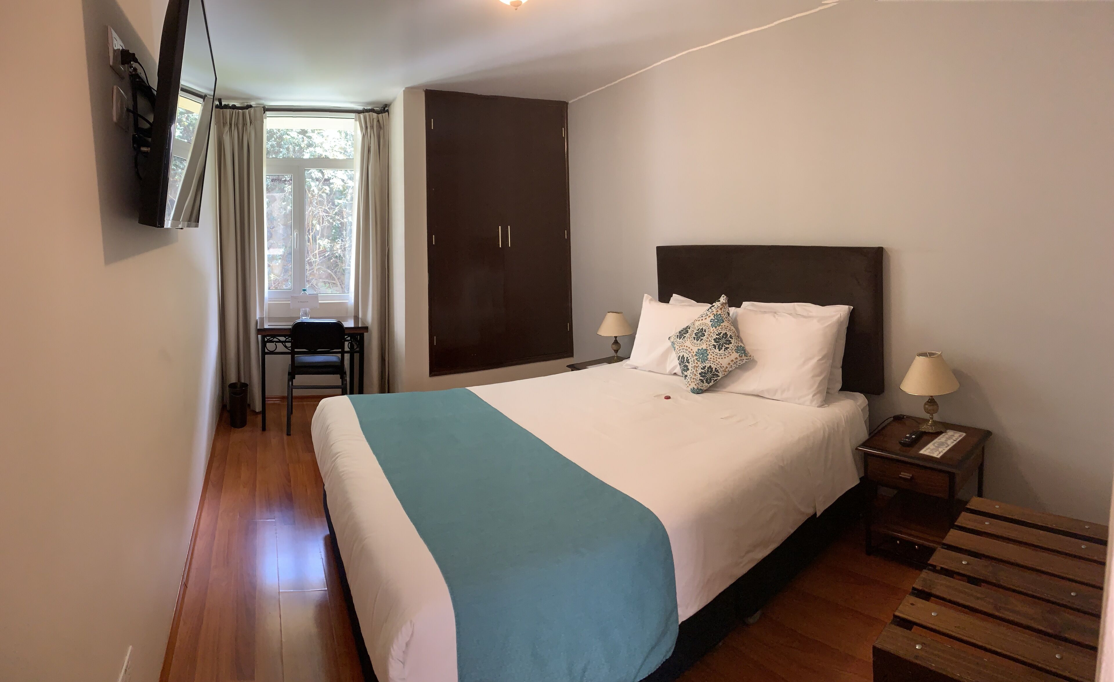 Quarto de hotel com cama, escrivaninha, cadeira, guarda-roupa e mesa de cabeceira com abajur.