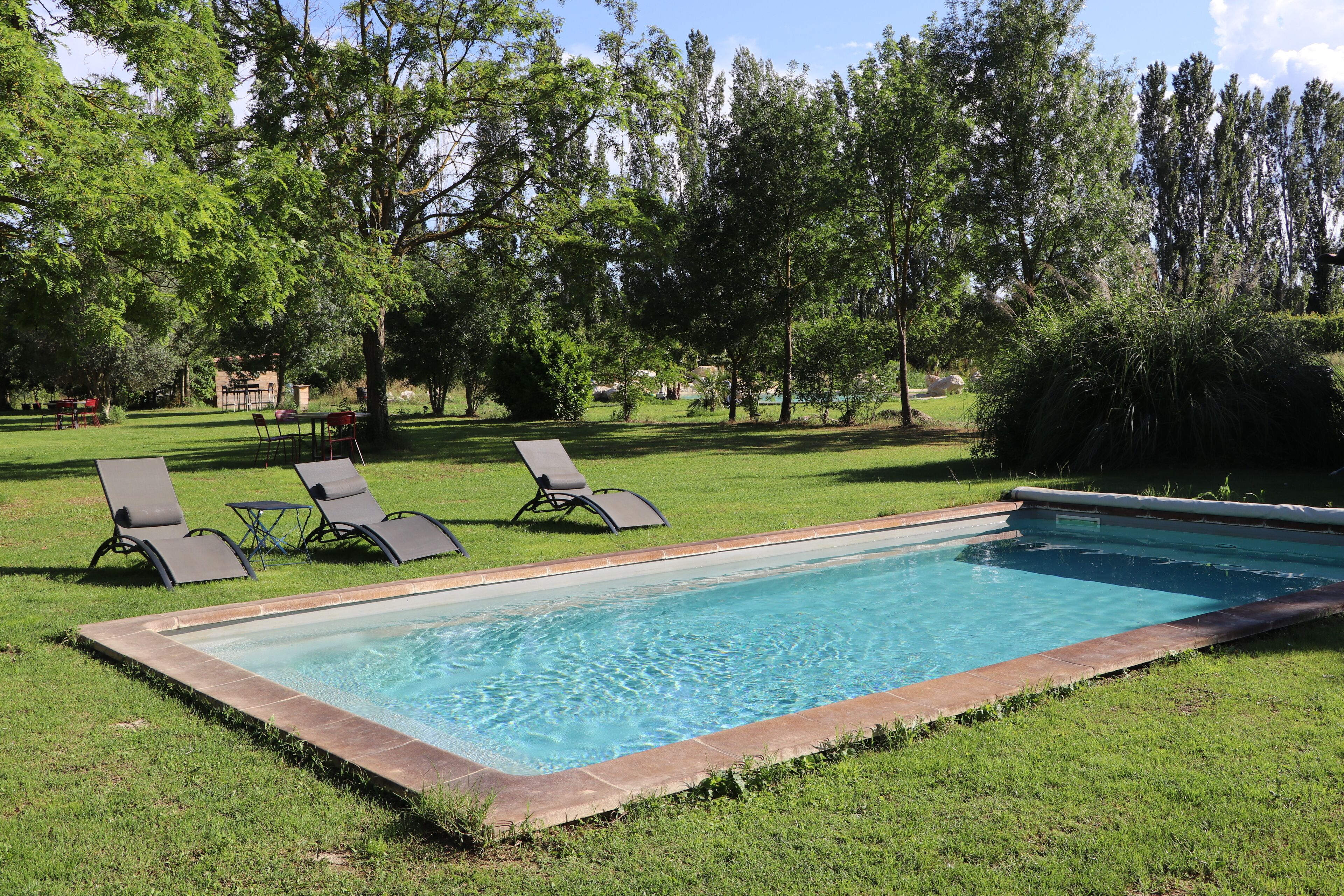Piscina retangular com borda azulejada, rodeada por uma área gramada com três espreguiçadeiras e uma mesa pequena.