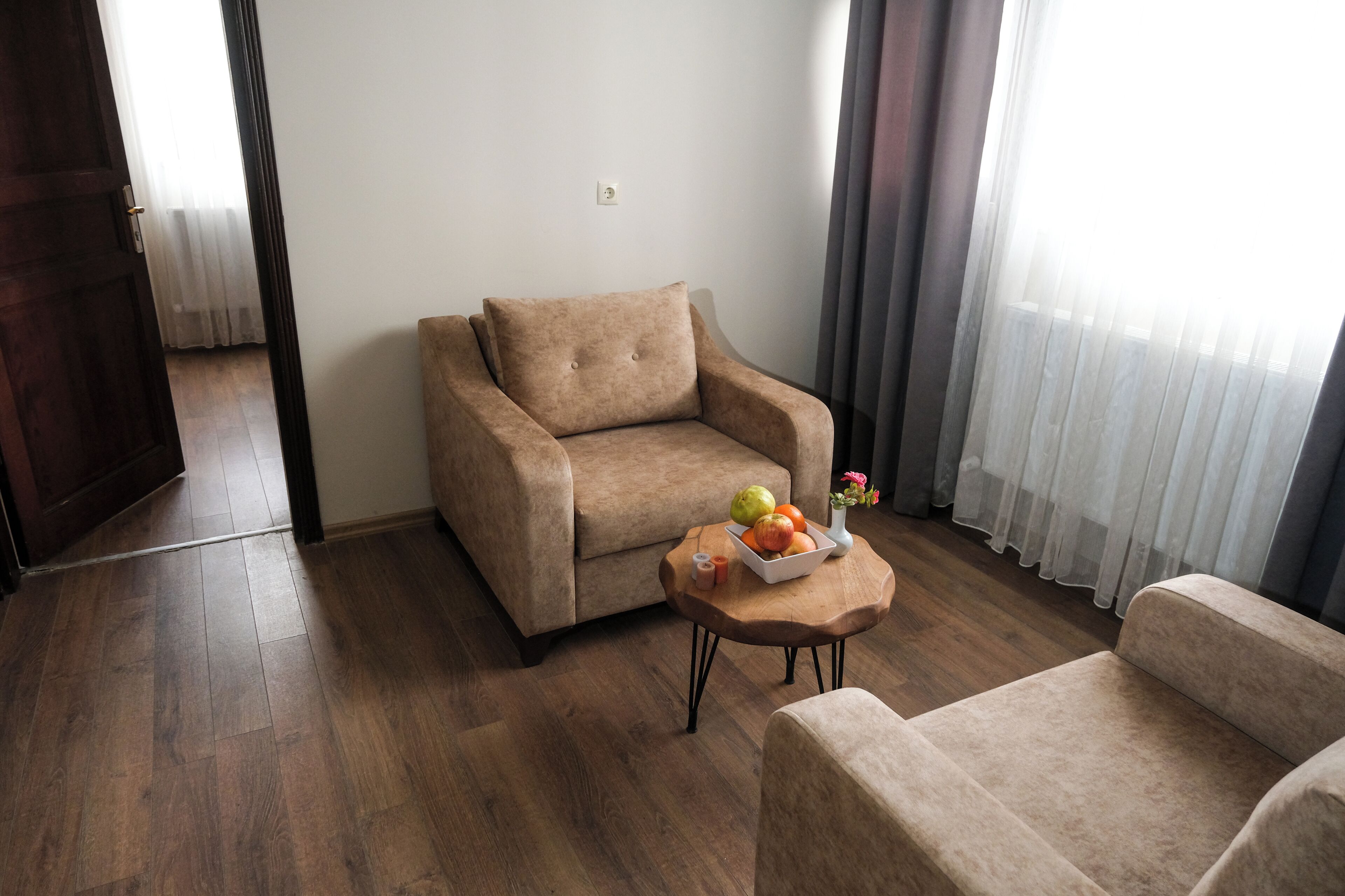 Sala de estar com um sofá bege, uma mesa de centro de madeira com frutas e um vaso, e vista através de cortinas translúcidas.
