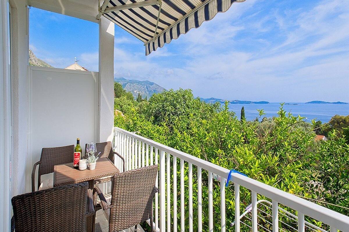 Uma varanda com móveis de vime, uma garrafa de vinho e vista para o mar e as montanhas.