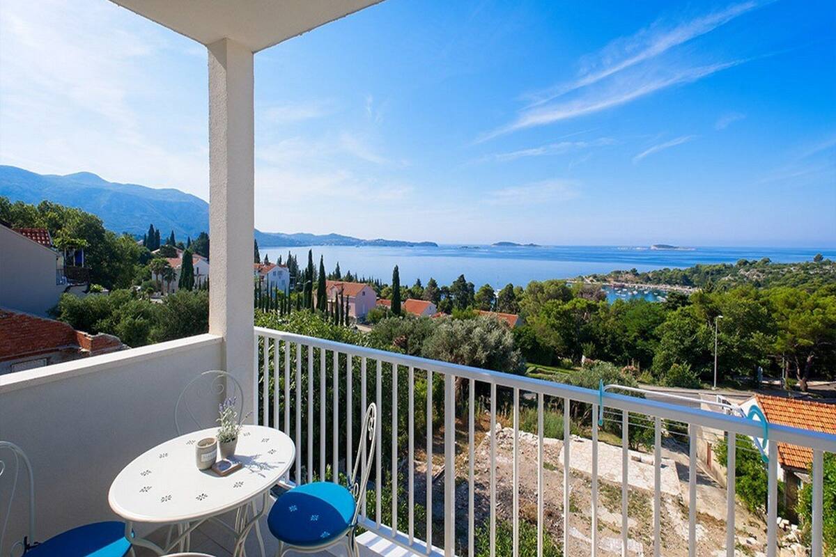 Uma varanda com mesa e cadeiras, com vista para uma paisagem costeira com casas e árvores.