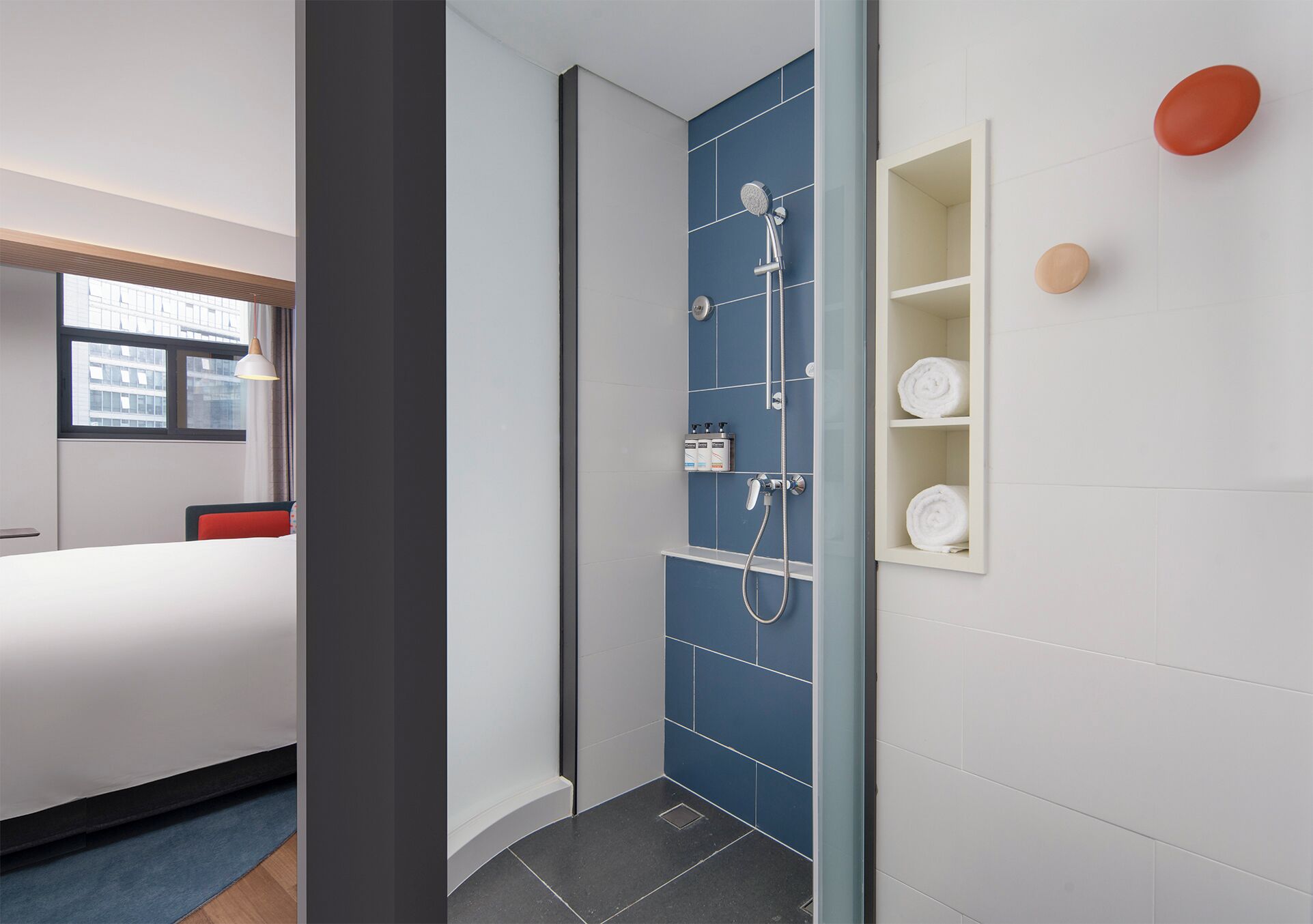 Quarto de hotel moderno com chuveiro, cama, janela e prateleira com toalhas.