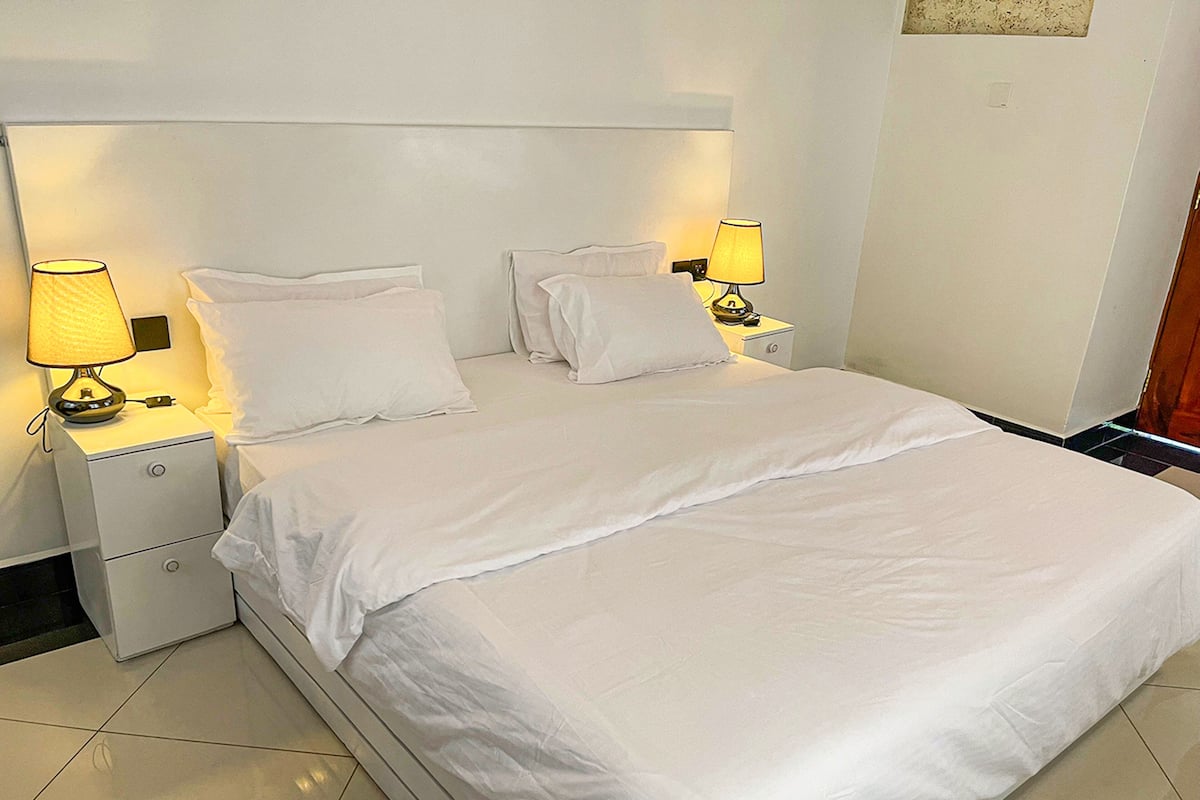 Cama bem arrumada com lençóis brancos, duas mesas de cabeceira com luminárias e uma parede lisa.