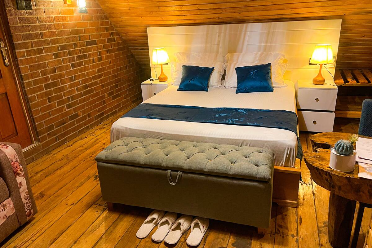 Quarto aconchegante com teto de madeira, parede de tijolos, cama com roupa de cama azul e branca, duas luminárias de cabeceira, mesa de cabeceira de madeira, um banco com chinelos e uma mesa de madeira com um cacto.