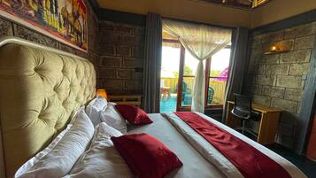 Cama com cabeceira, roupa de cama branca e almofadas vermelhas. Há uma luminária, uma cadeira e uma televisão sobre uma mesa de madeira. O quarto tem paredes de pedra e um quadro na parede.