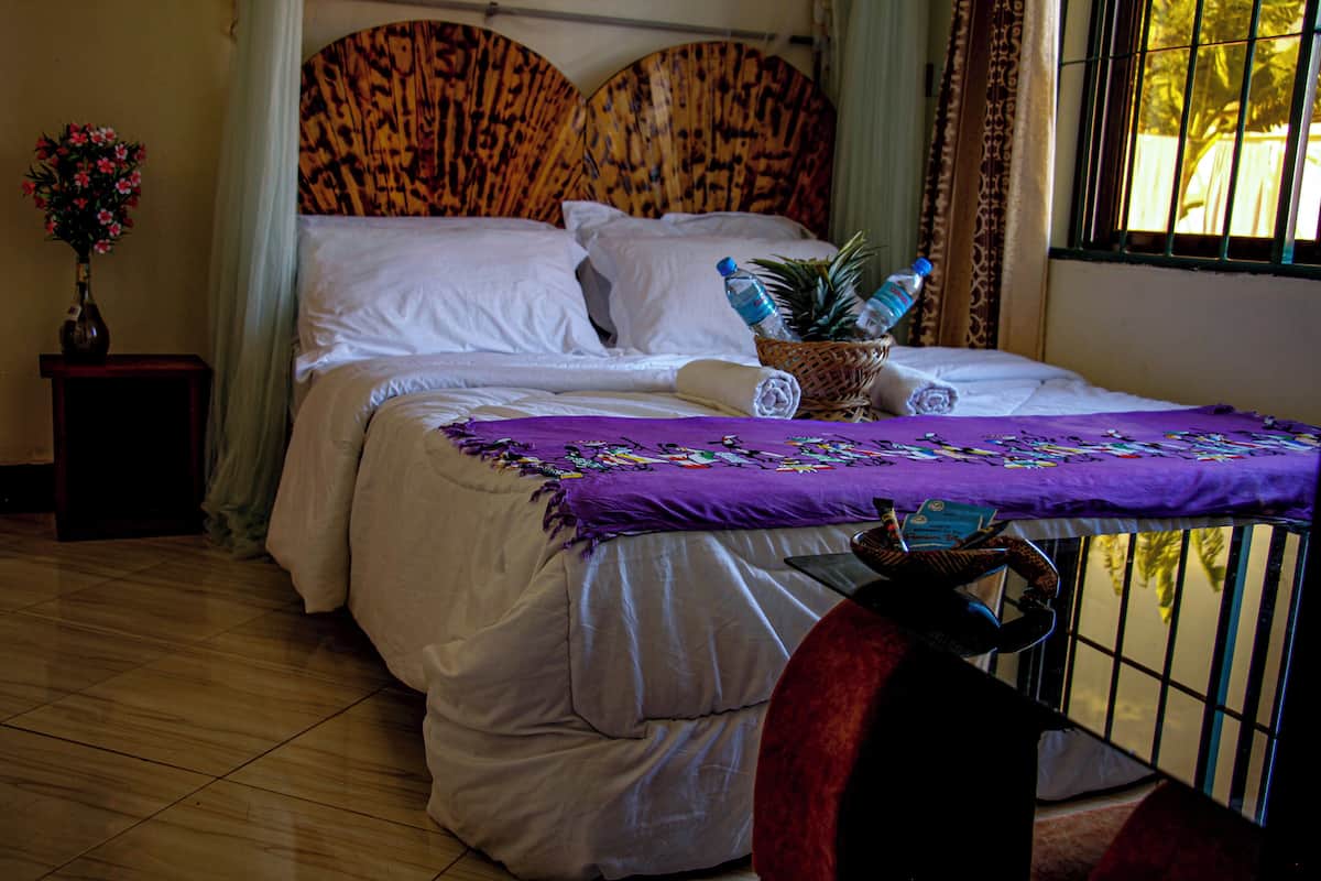 Uma cama com um cobertor roxo, almofadas brancas e uma cesta de palha com uma garrafa e uma planta.