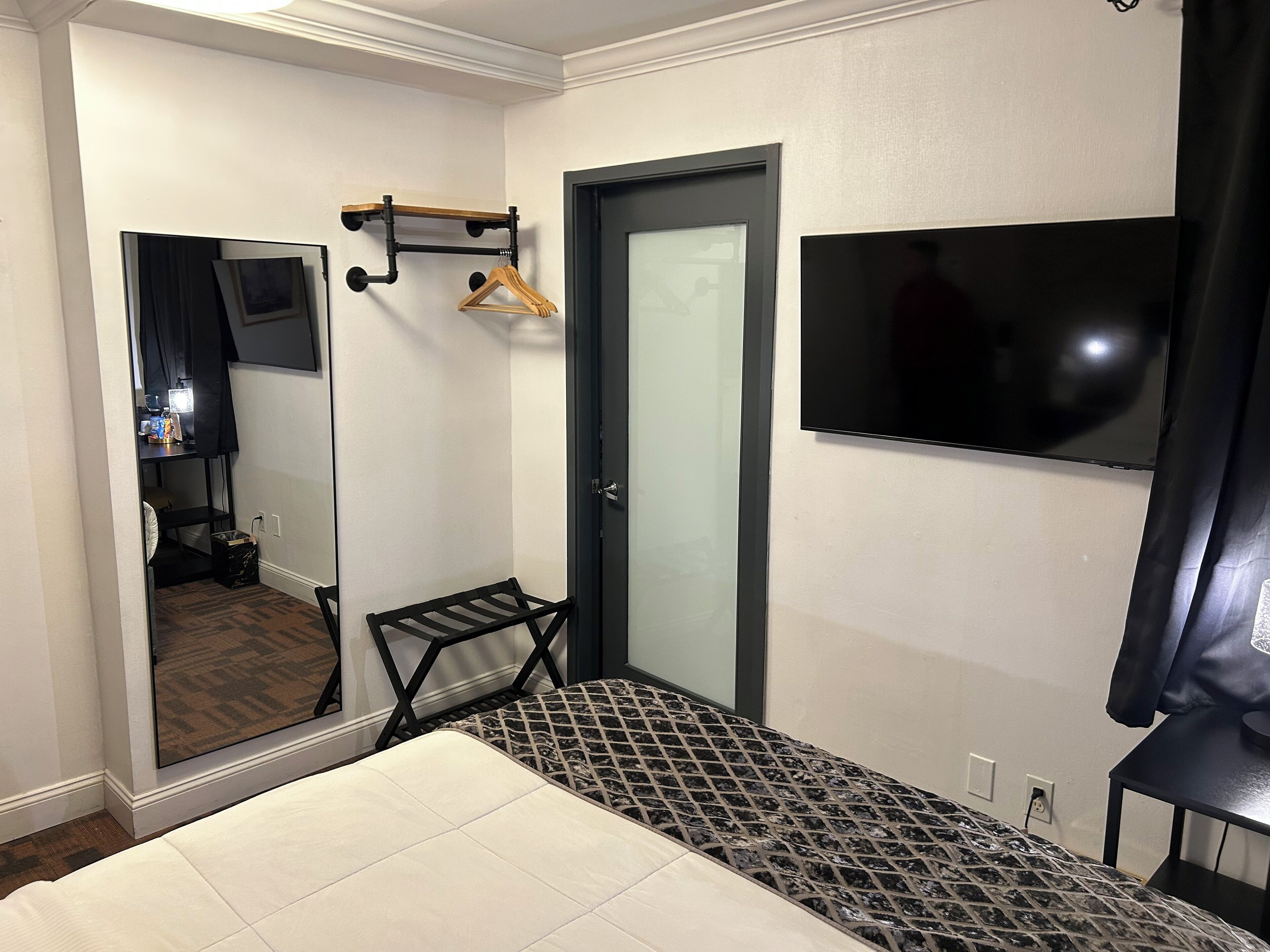 Quarto de hotel com cama, televisão na parede, espelho e um pequeno espaço de estar com um banco.