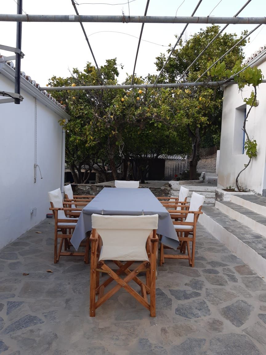Uma área de refeições ao ar livre coberta, com uma mesa e cadeiras, cercada por um muro branco e árvores.