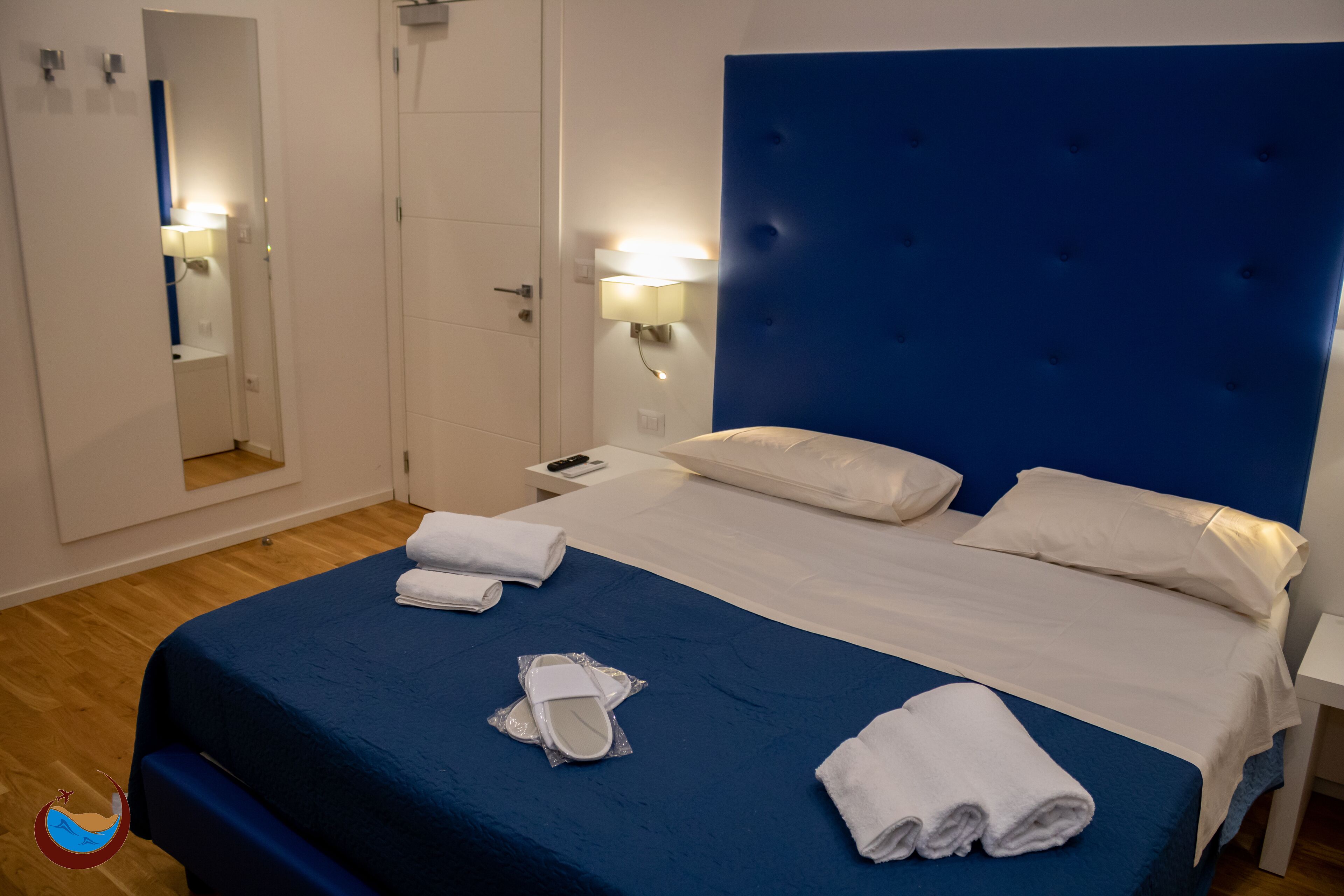 Quarto de hotel com uma cama grande, cabeceira azul, almofadas brancas e mesa de cabeceira com abajur.