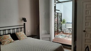 Quarto com cama, almofadas, mesa de cabeceira e porta que dá para uma varanda com mesa e cadeiras.