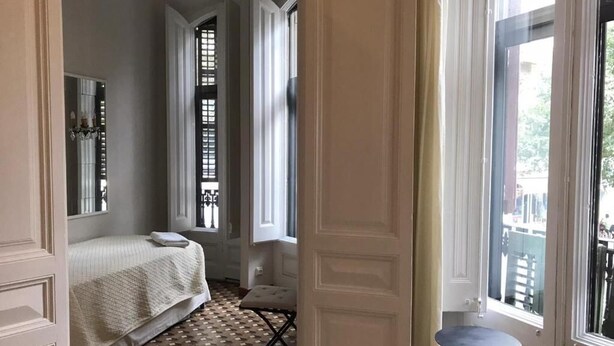Quarto com cama, uma mesinha e uma cadeira. Janelas com persianas e vista para o exterior.