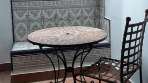 Uma pequena área de pátio fechada com uma mesa redonda e uma única cadeira. As paredes são revestidas com um padrão decorativo, e o chão com azulejos de terracota.