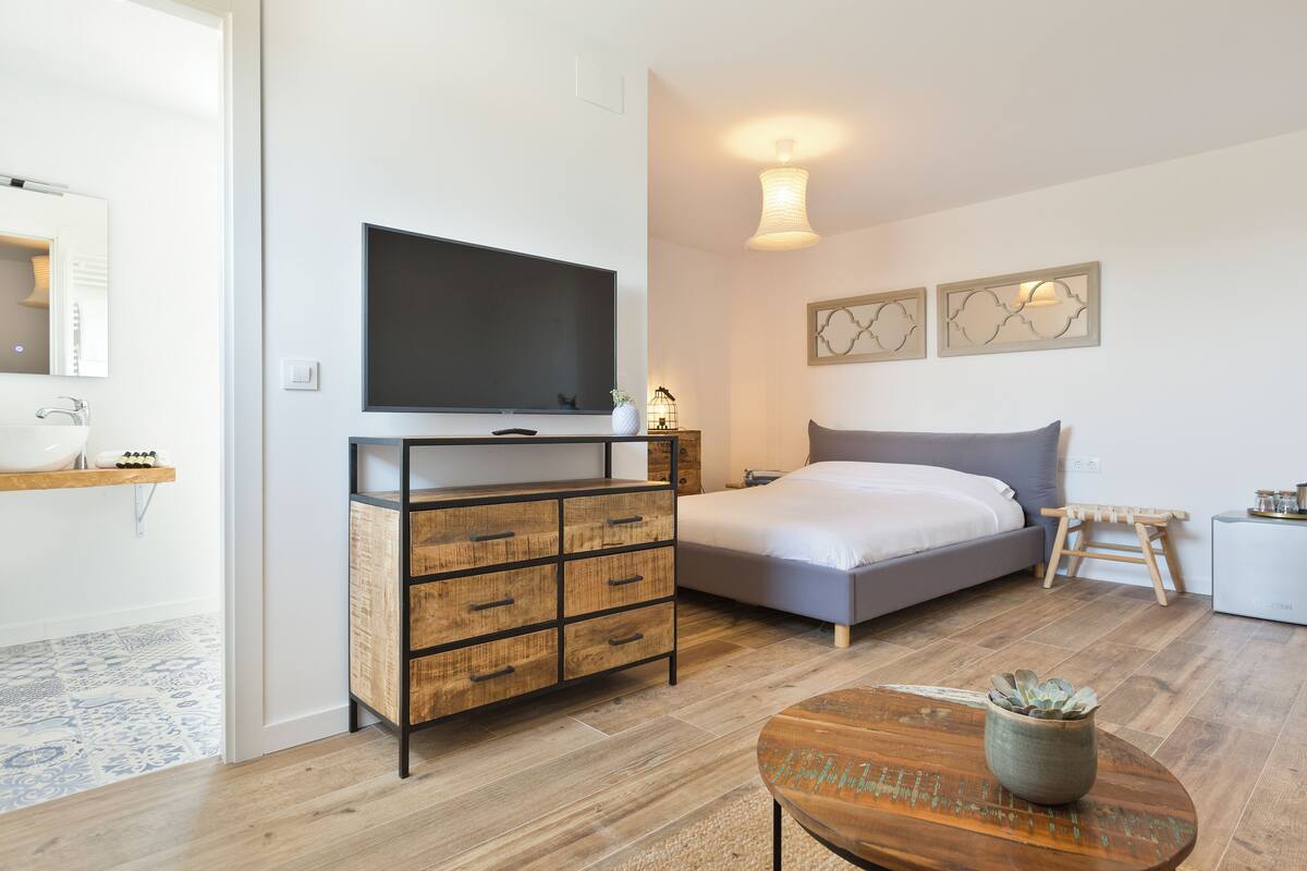Quarto moderno com cama, televisão, cómoda de madeira e uma pequena mesa com uma planta.