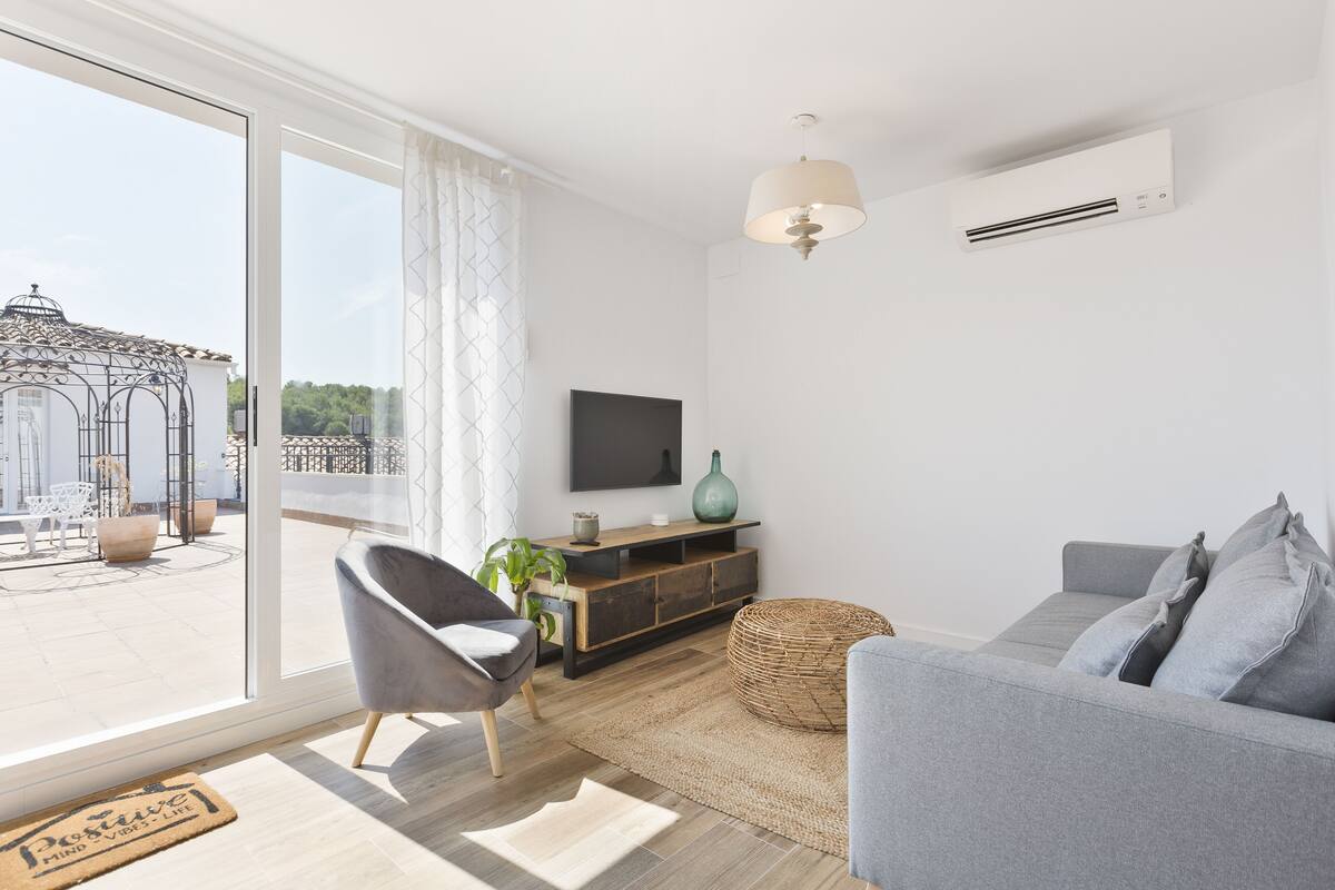 Uma sala de estar moderna com um sofá cinzento, uma poltrona preta, um móvel de TV em madeira e uma grande janela com vista para um pátio com mobiliário de exterior.