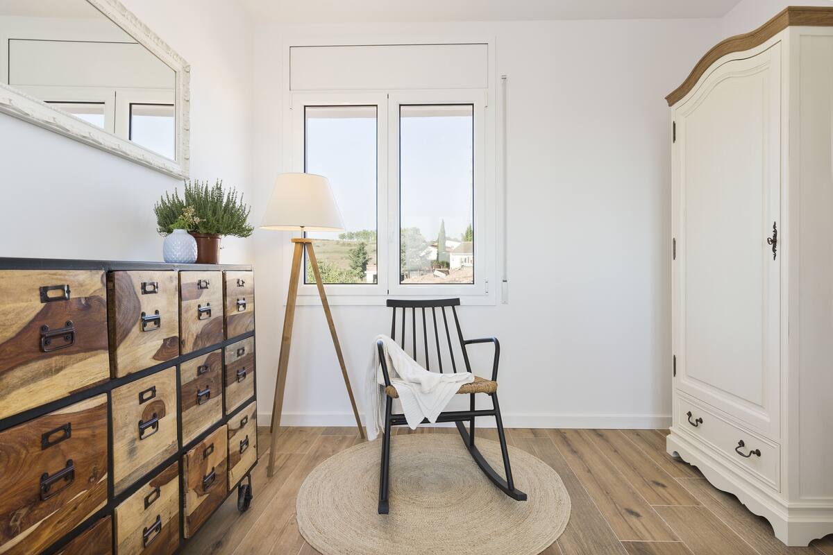 Quarto com penteadeira de madeira, uma cadeira de balanço, luminária e vista pela janela.