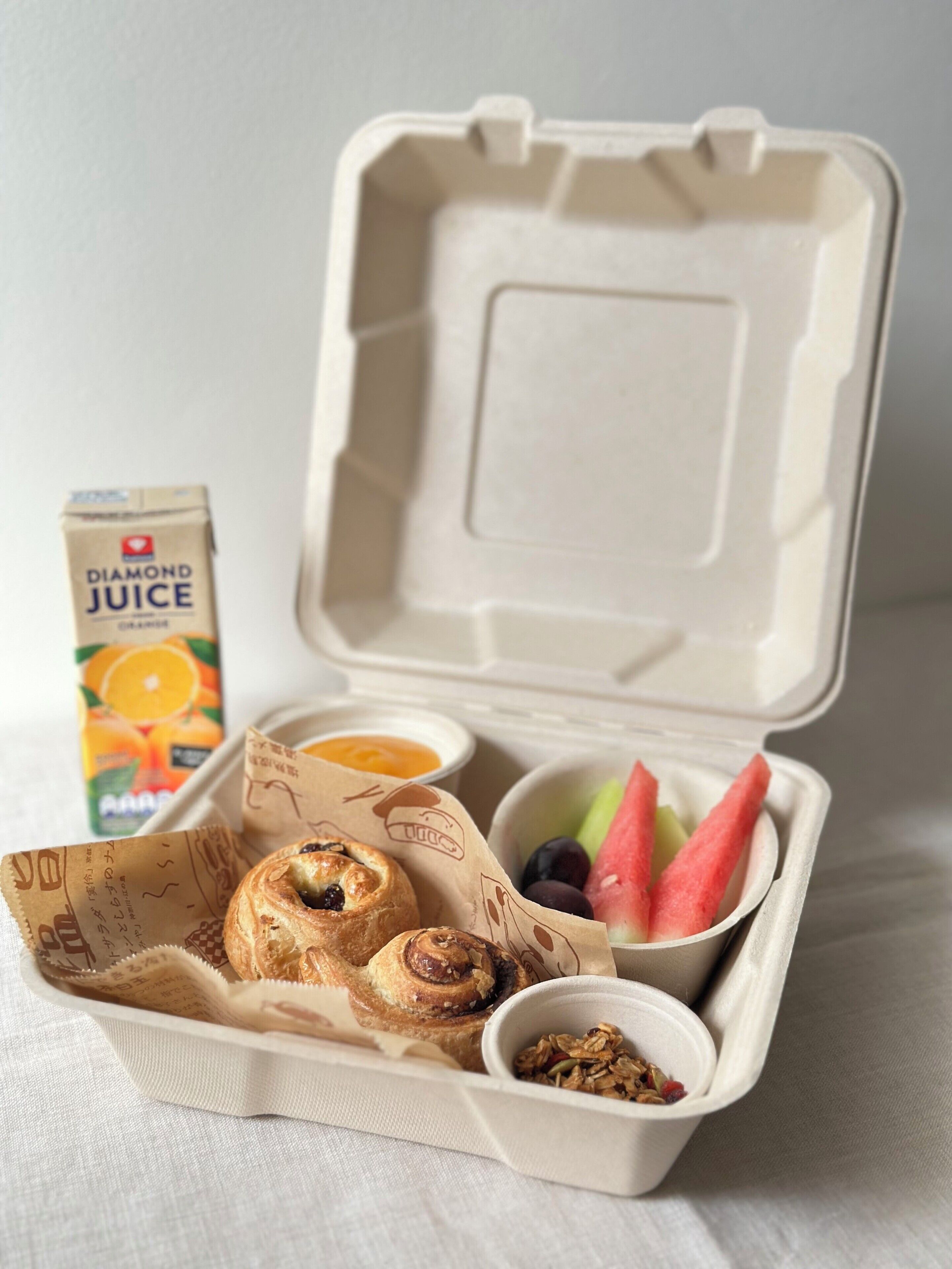 Recipiente de comida com suco em cartão, croissants, melancia, uvas e granola.