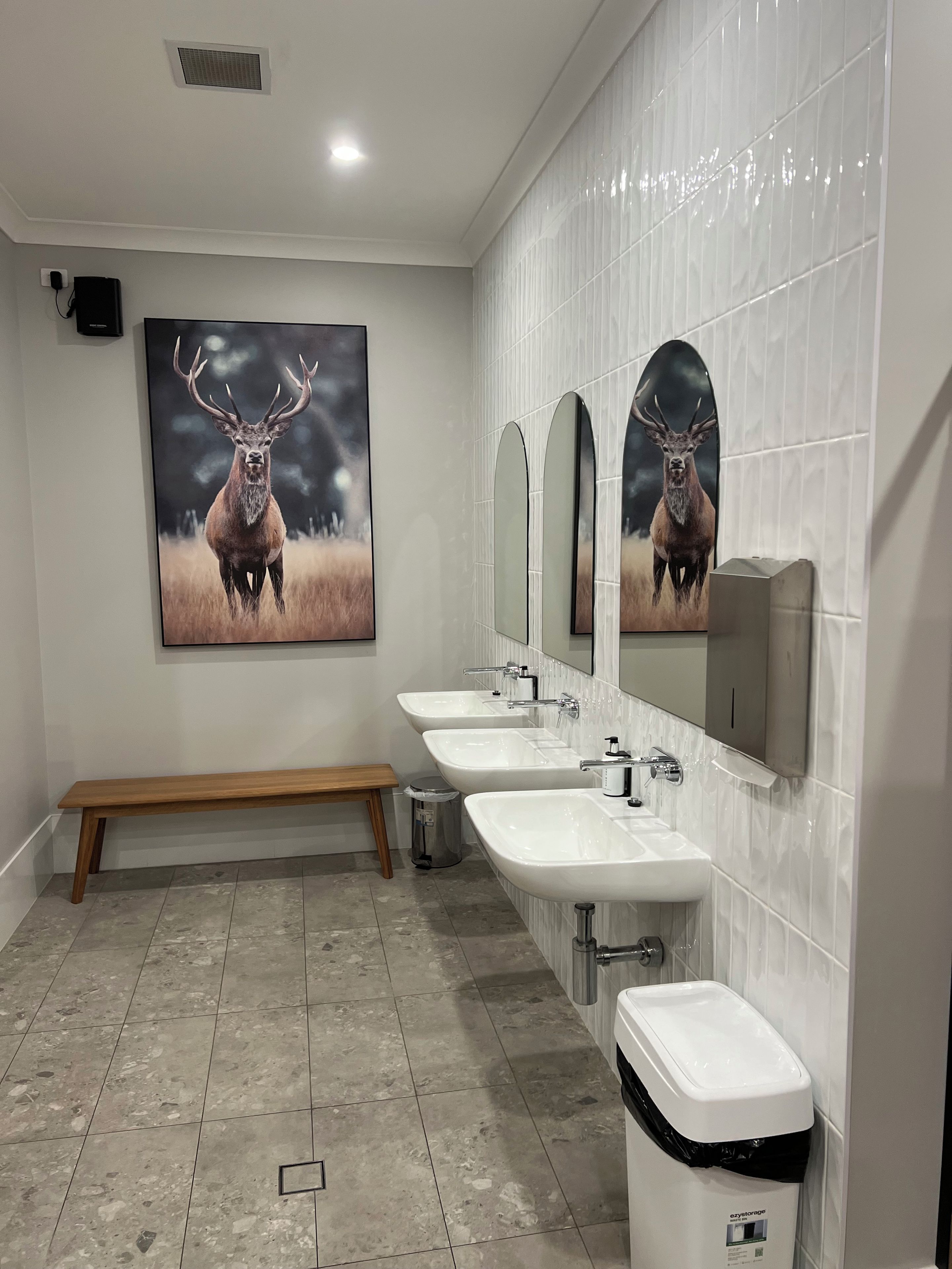 Casa de banho com três pias, um banco e um cestinho de lixo.