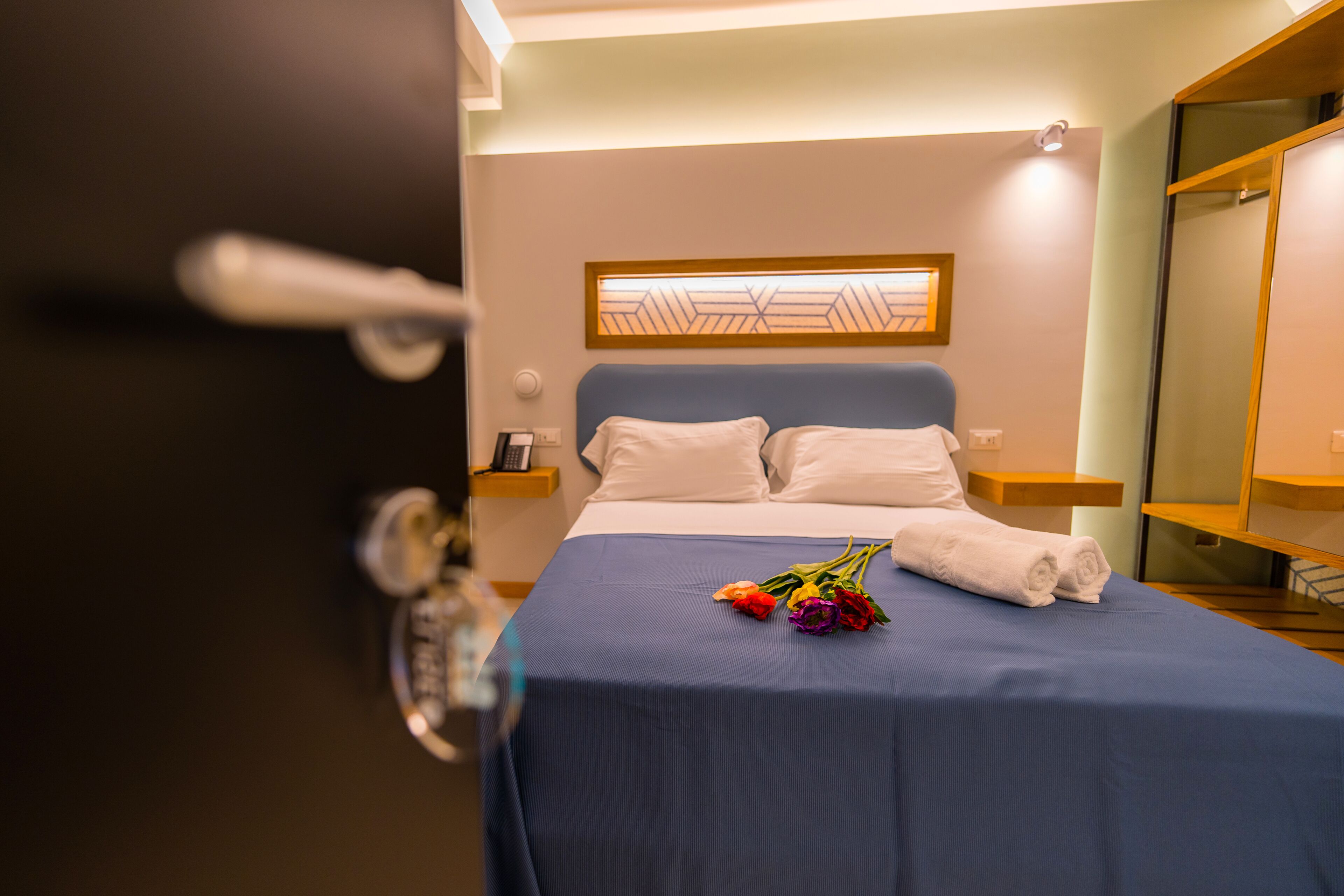 Quarto de hotel com cama bem arrumada, mesa de cabeceira com luminária e janela com padrão geométrico.