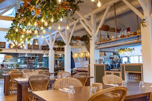 Um restaurante com mesas de madeira e cadeiras de vime, luminárias pendentes e uma cozinha ao fundo.