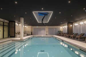 Piscina interior moderna com luminária geométrica sobre a água, cadeiras de descanso ao longo da borda e vista do céu através de uma janela.