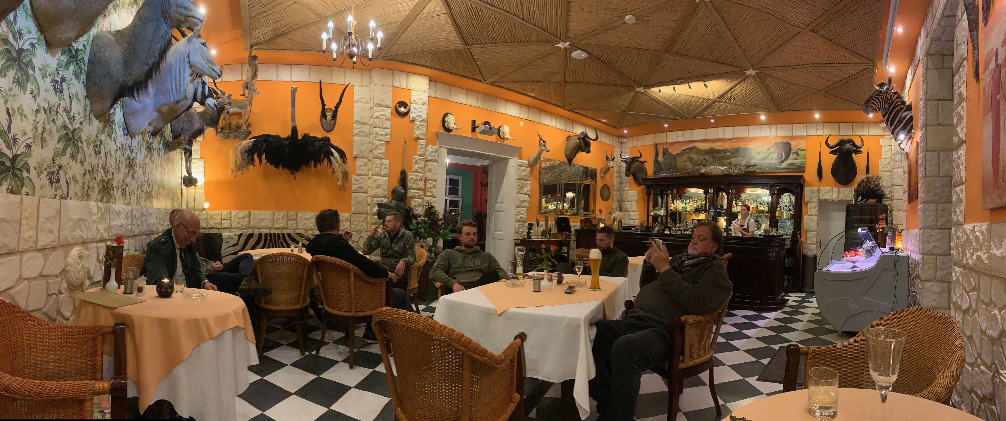 Um restaurante com chão xadrez, mesas com toalhas brancas e várias decorações de cabeça de animais nas paredes.