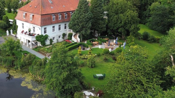 Um edifício grande e branco com telhado de telhas vermelhas, rodeado por vegetação exuberante e um lago.