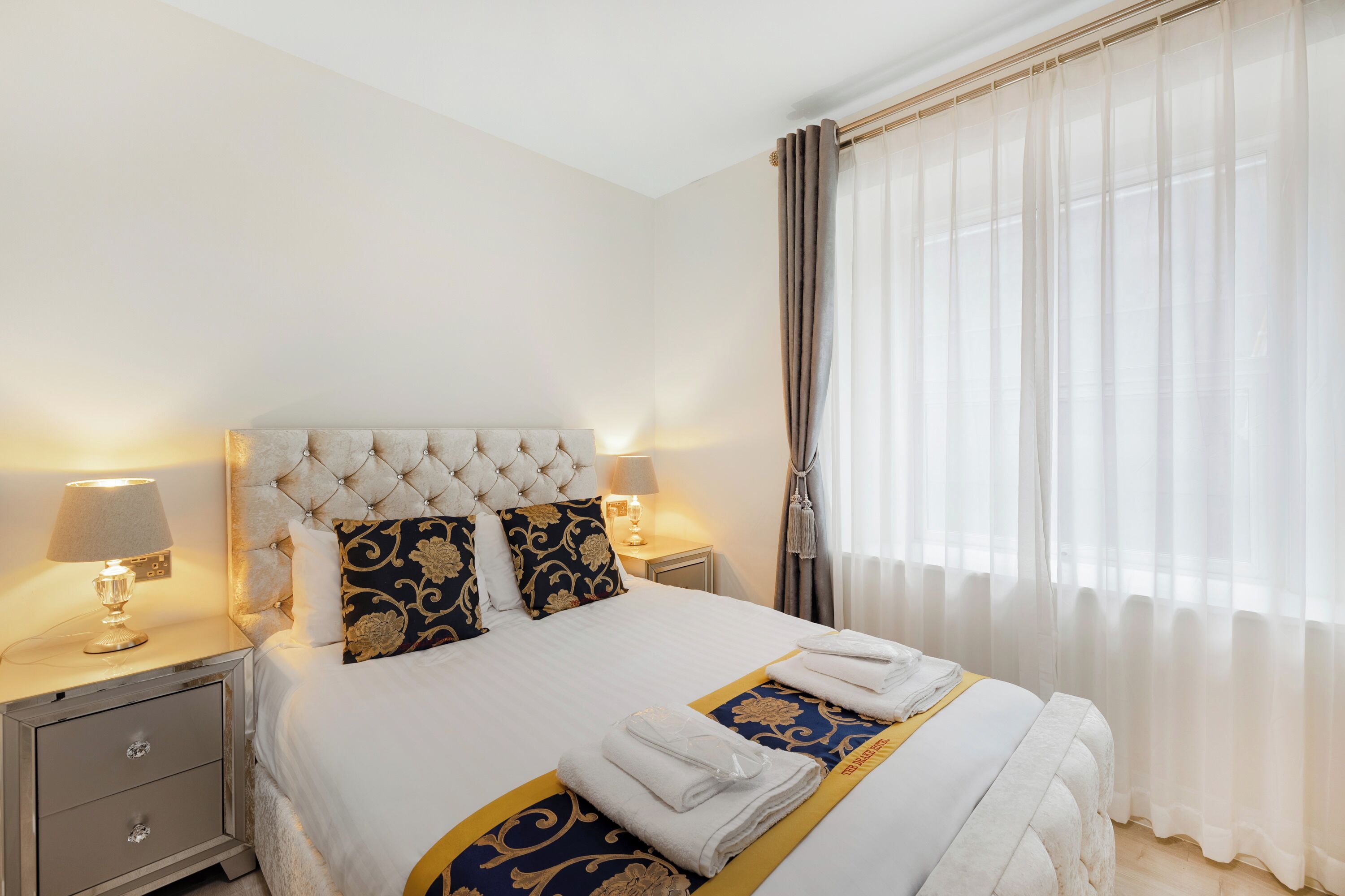 Quarto de hotel com uma cama grande, duas mesas de cabeceira com luminárias, uma janela com cortinas leves e uma cama bem arrumada com toalhas.