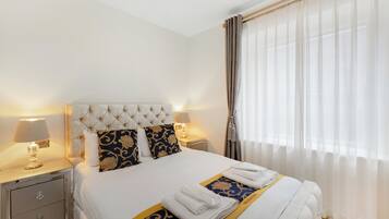 Quarto de hotel com uma cama grande, duas mesas de cabeceira com luminárias, uma janela com cortinas leves e uma cama bem arrumada com toalhas.