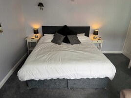 Uma cama bem arrumada com roupa de cama branca e duas almofadas decorativas, acompanhada por mesas de cabeceira com abajures, em um quarto com cabeceira escura e paredes claras.