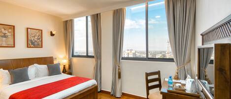 Quarto de hotel com uma cama grande, uma escrivaninha com cadeira e vista para a cidade através de amplas janelas.