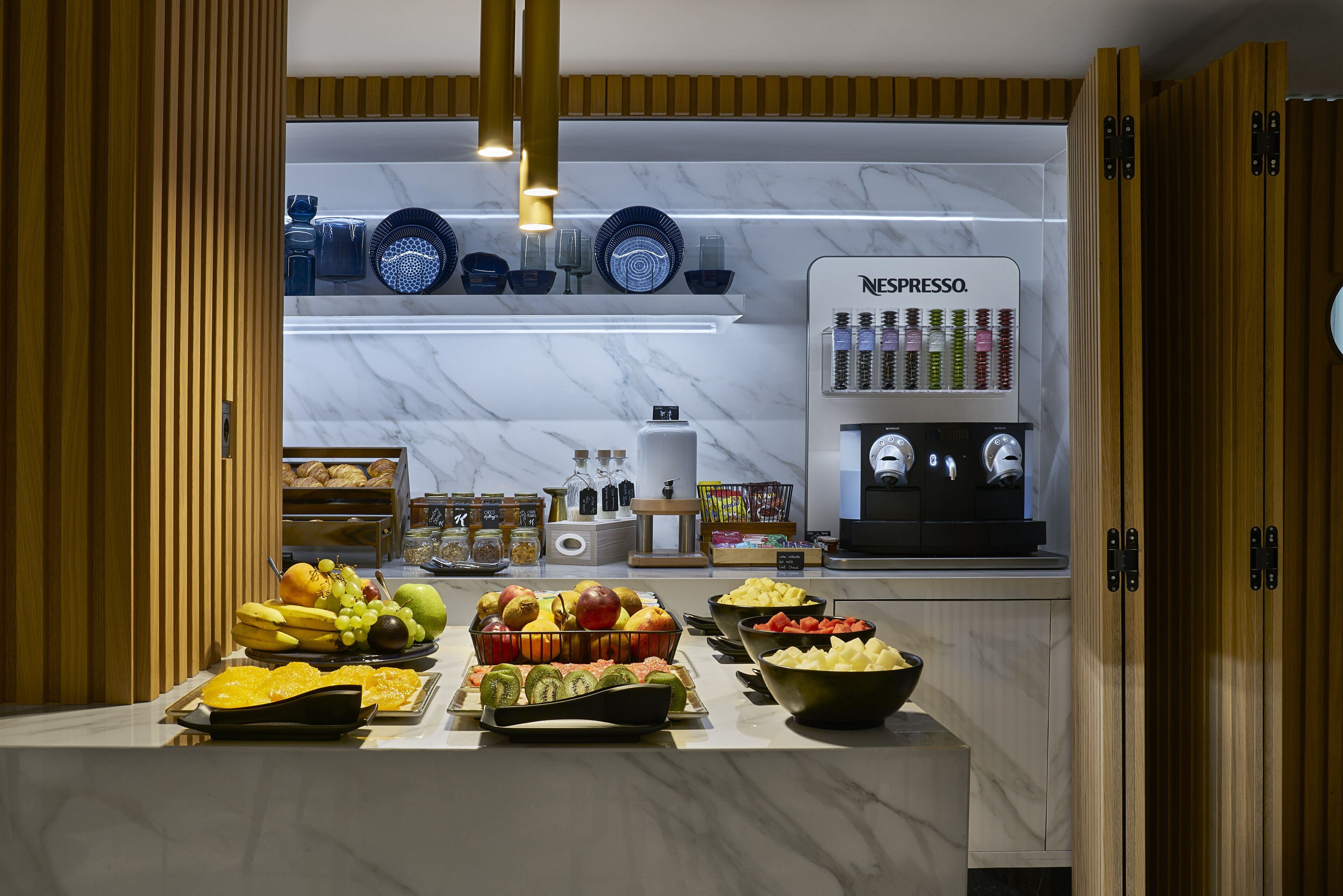 Um buffet de café da manhã com diversas frutas e doces, um espaço para café com máquinas Nespresso e um painel de fundo de mármore.