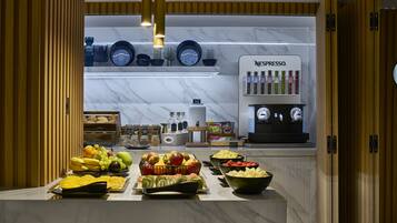 Um buffet de café da manhã com diversas frutas e doces, um espaço para café com máquinas Nespresso e um painel de fundo de mármore.