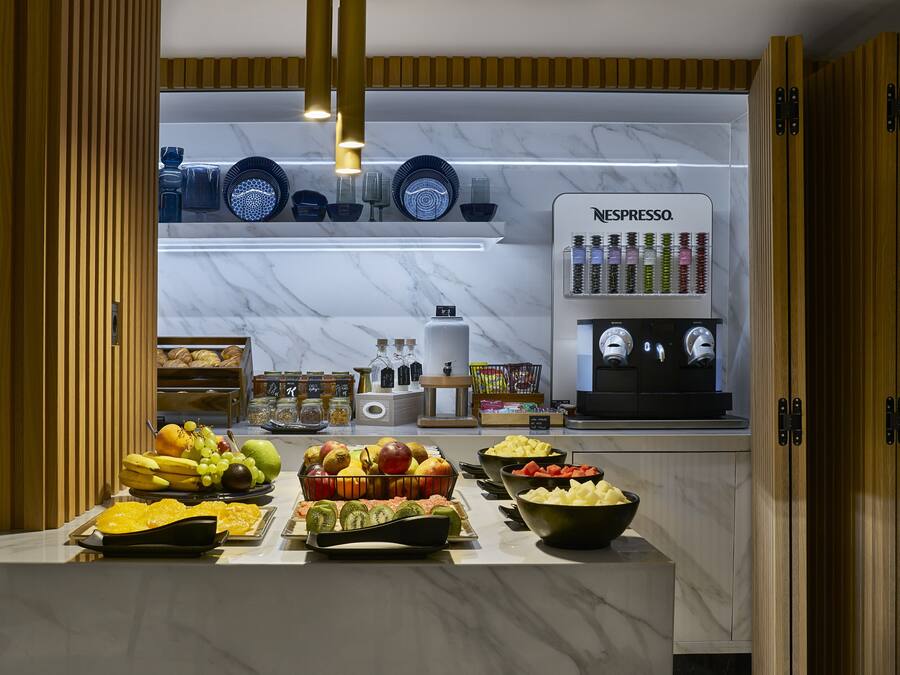 Um buffet de café da manhã com diversas frutas e doces, um espaço para café com máquinas Nespresso e um painel de fundo de mármore.