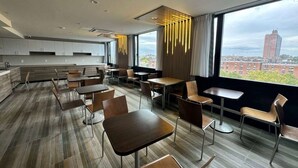 Uma área de refeições moderna com várias mesas e cadeiras, grandes janelas com vista para a cidade e uma cozinha ao fundo.