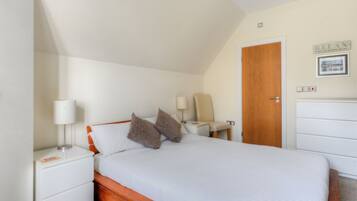Quarto com cama de madeira, mesas de cabeceira brancas, porta de madeira e cômoda branca.