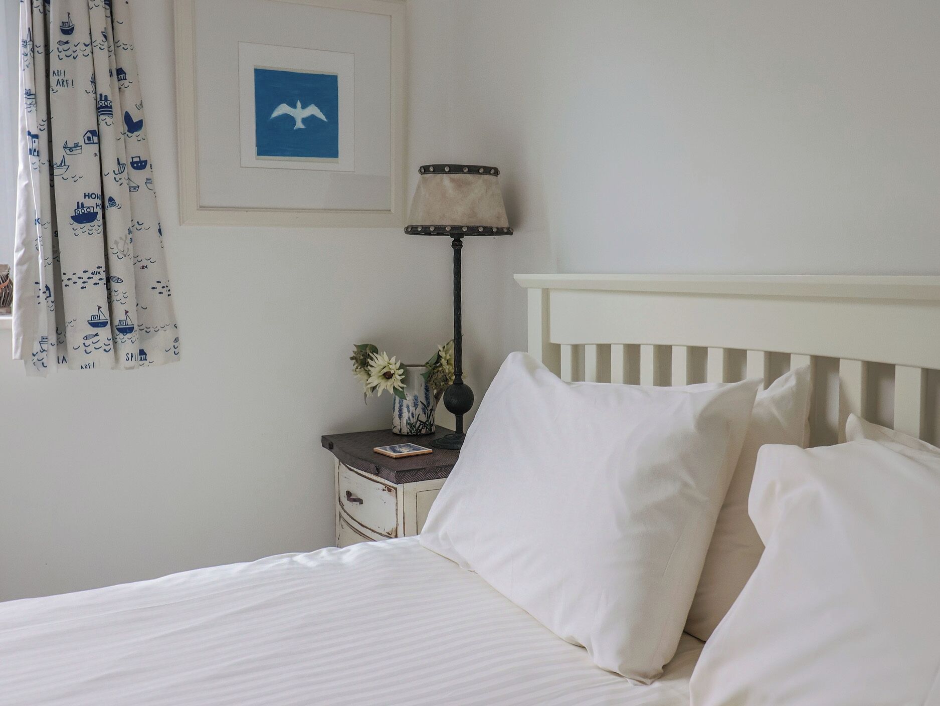 Uma cama bem arrumada com roupa de cama branca, uma mesa de cabeceira com abajur, um quadro na parede e cortinas com estampas náuticas.