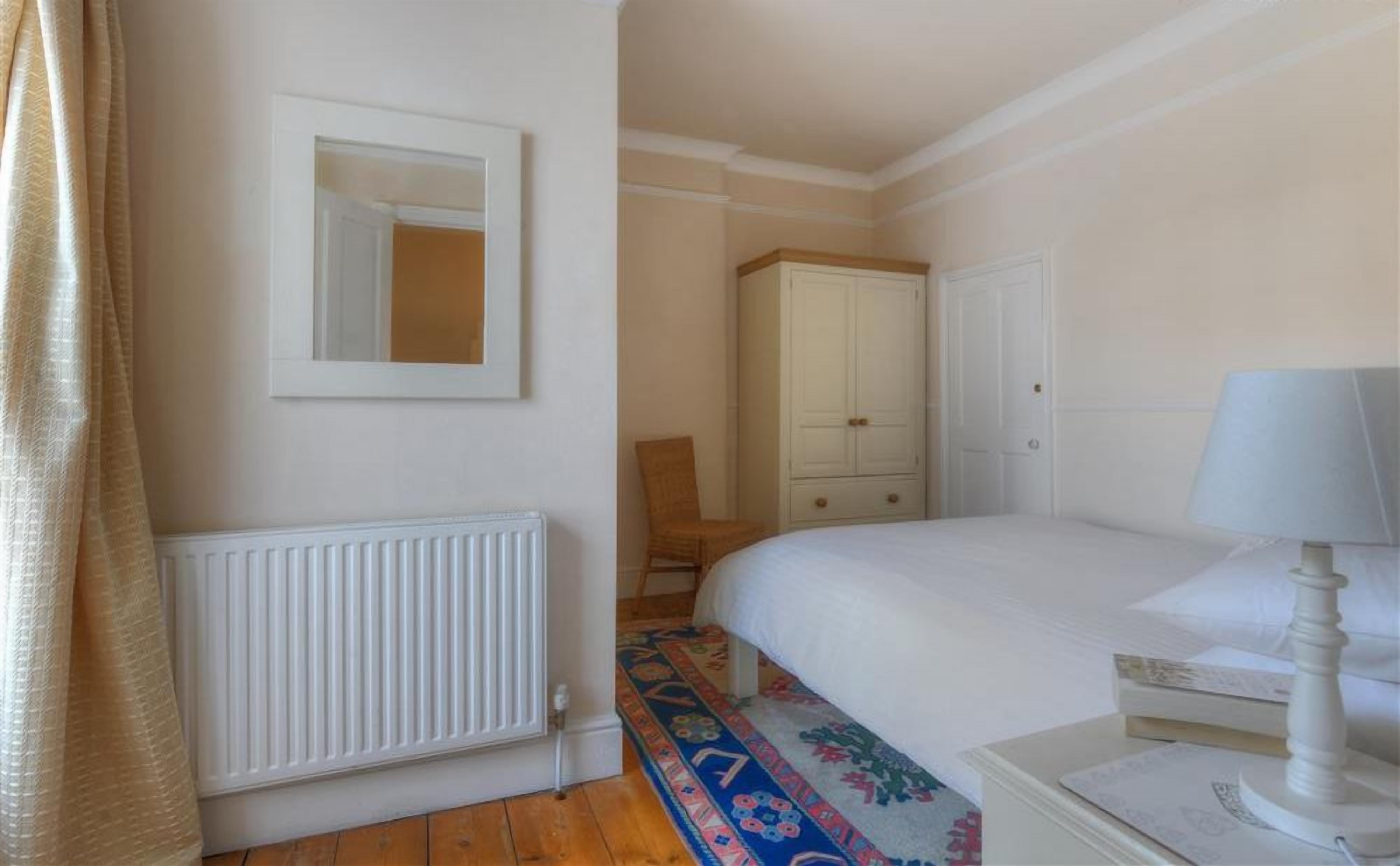 Um quarto com cama, mesa de cabeceira com abajur, guarda-roupa, radiador e espelho na parede.