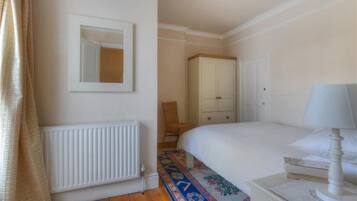 Um quarto com cama, mesa de cabeceira com abajur, guarda-roupa, radiador e espelho na parede.