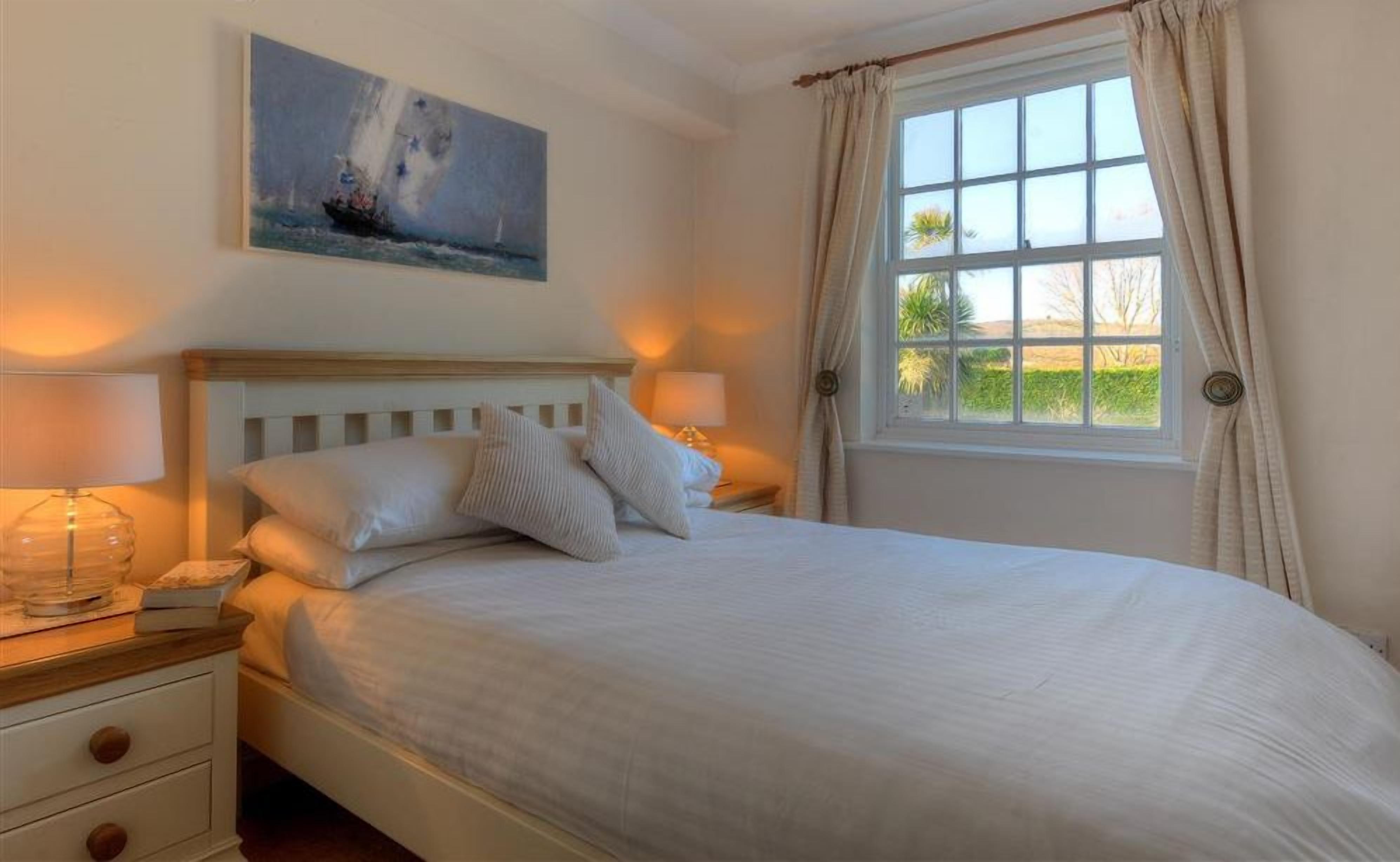 Quarto com uma cama grande, duas mesas de cabeceira, um criado-mudo, janela com cortinas e um quadro na parede.