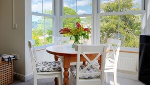 Quarto com mesa redonda e cadeiras brancas, um vaso com flores e grandes janelas que dão para árvores e edifícios.