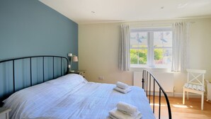 Um quarto com cama, mesa de cabeceira, luminária, janela com cortinas, radiador e uma cadeira.