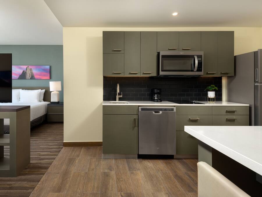 Quarto de hotel compacto com cozinha compacta, cama com roupa de cama branca, mesa de cabeceira e televisão presa à parede.