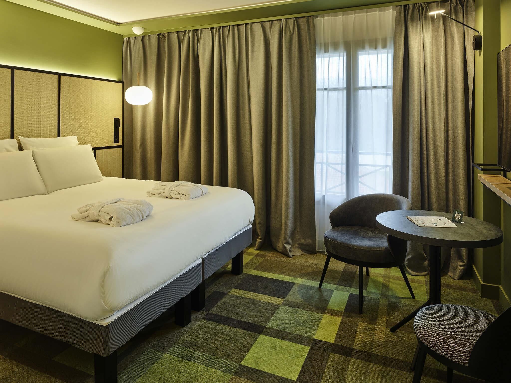Quarto de hotel com cama, uma cadeira, uma mesinha e vista para o exterior pela janela.