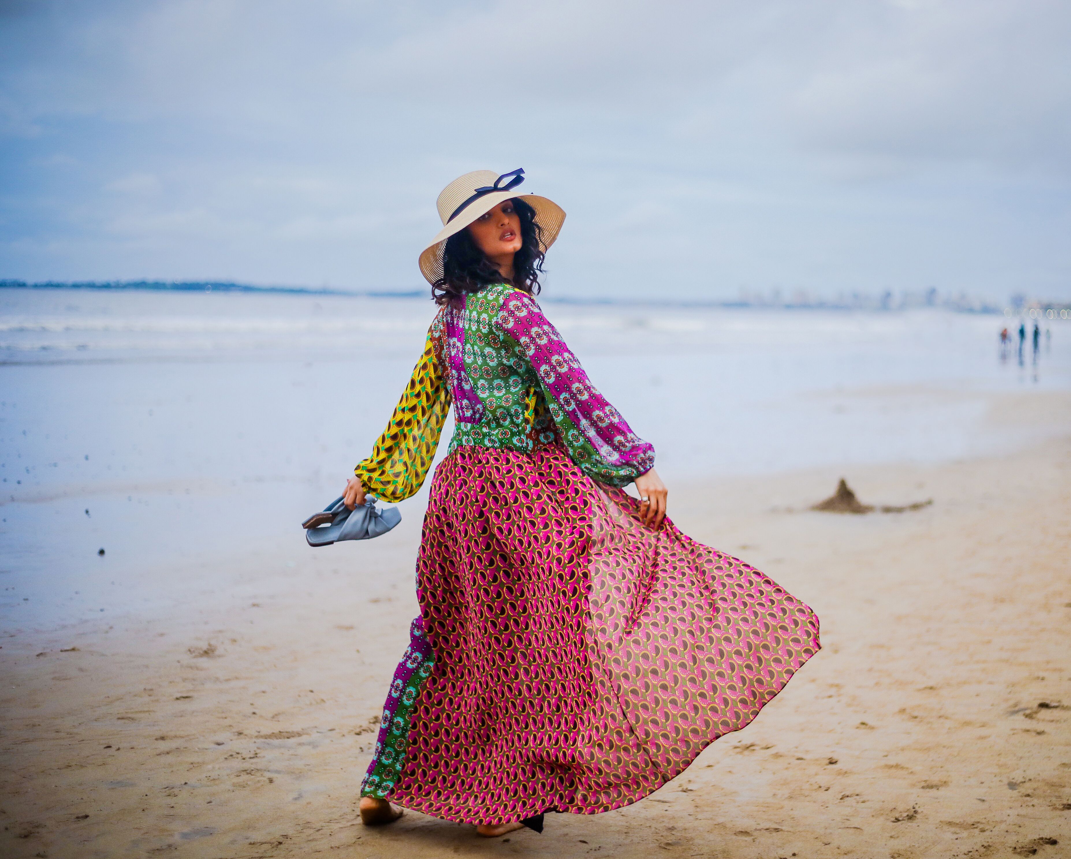 Uma pessoa numa praia arenosa, vestindo um vestido estampado e um chapéu, segurando um sapato.