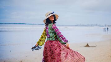 Uma pessoa numa praia arenosa, vestindo um vestido estampado e um chapéu, segurando um sapato.