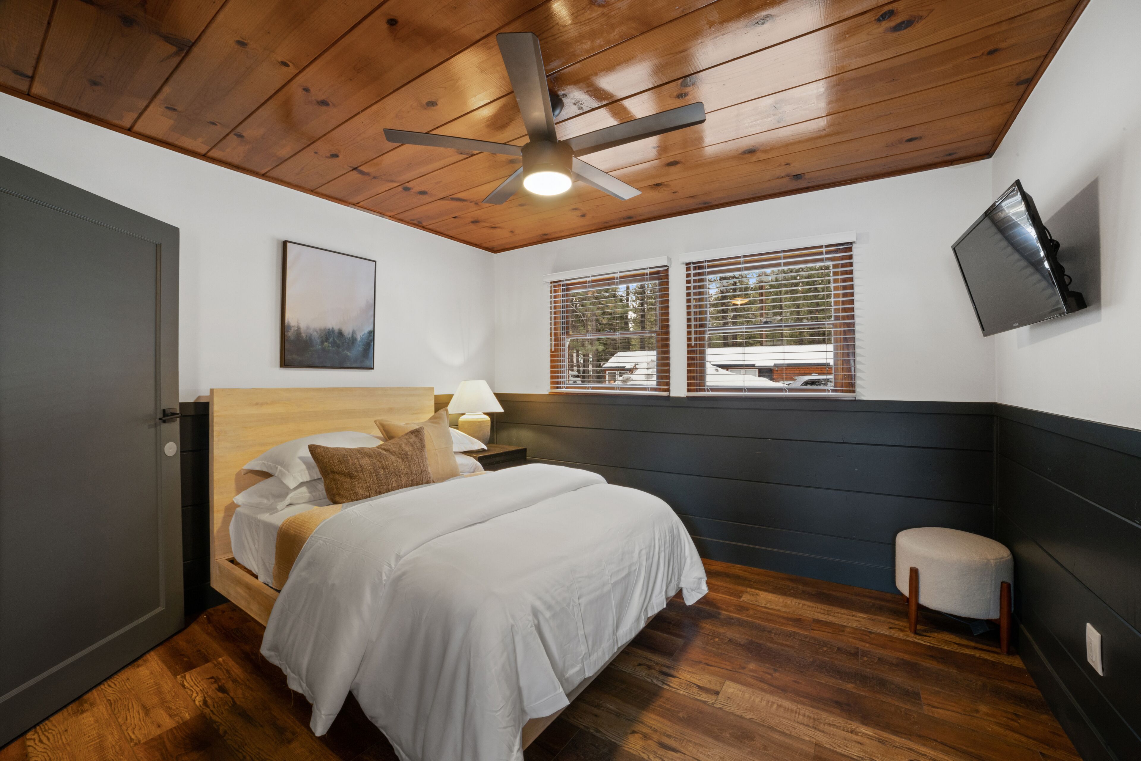 Quarto com teto de madeira, cama com roupa de cama branca, mesa de cabeceira com luminária e TV fixada na parede.