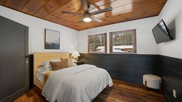 Quarto com teto de madeira, cama com roupa de cama branca, mesa de cabeceira com luminária e TV fixada na parede.