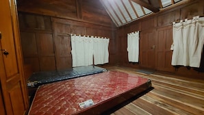 Quarto com paredes de madeira e teto inclinado. Há duas camas com colchões, uma vermelha e outra preta. Cortinas brancas adornam as janelas.