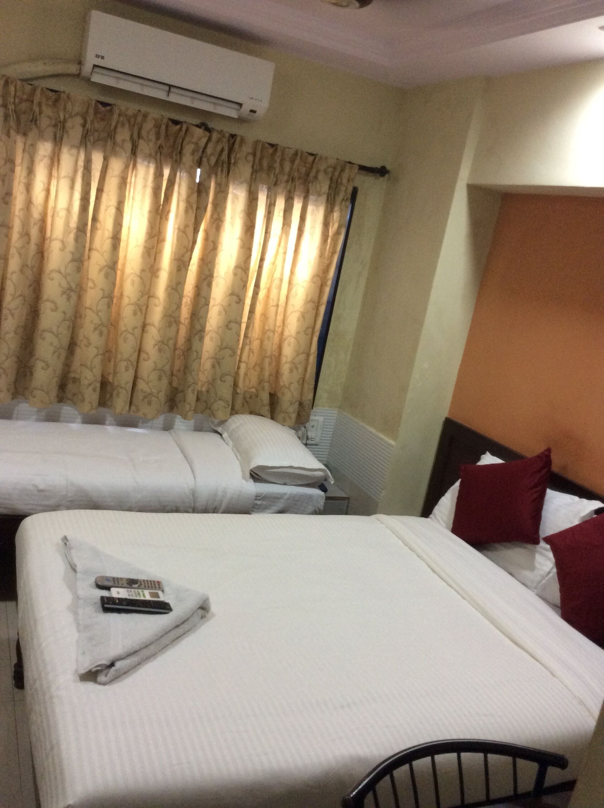 Quarto de hotel com duas camas, uma janela com cortinas, um ar condicionado e uma mesa de cabeceira com controle remoto.