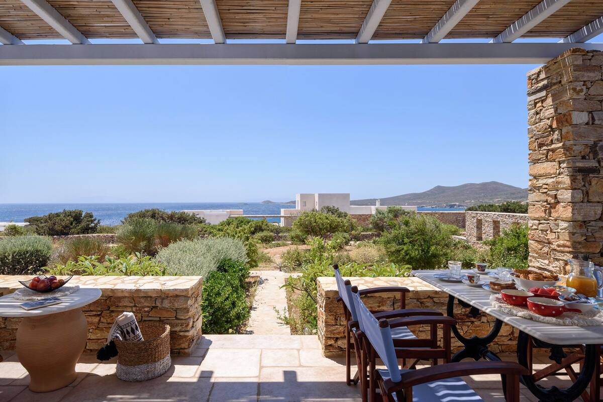 Uma área de refeições ao ar livre com pilares de pedra, uma mesa posta para o café da manhã e vista para o mar e colinas.